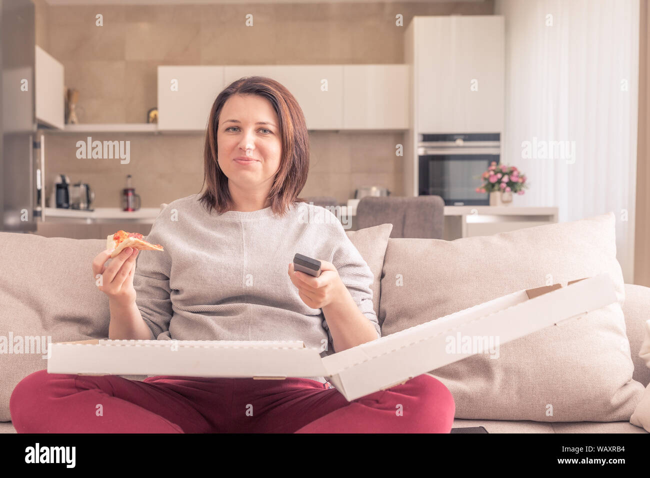 Beautiful woman eating pizza and watching TV holding remote control at ...