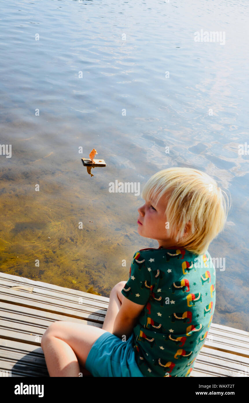 Young blonde boy sitting on jetty with wooden toy boat on lake in ...