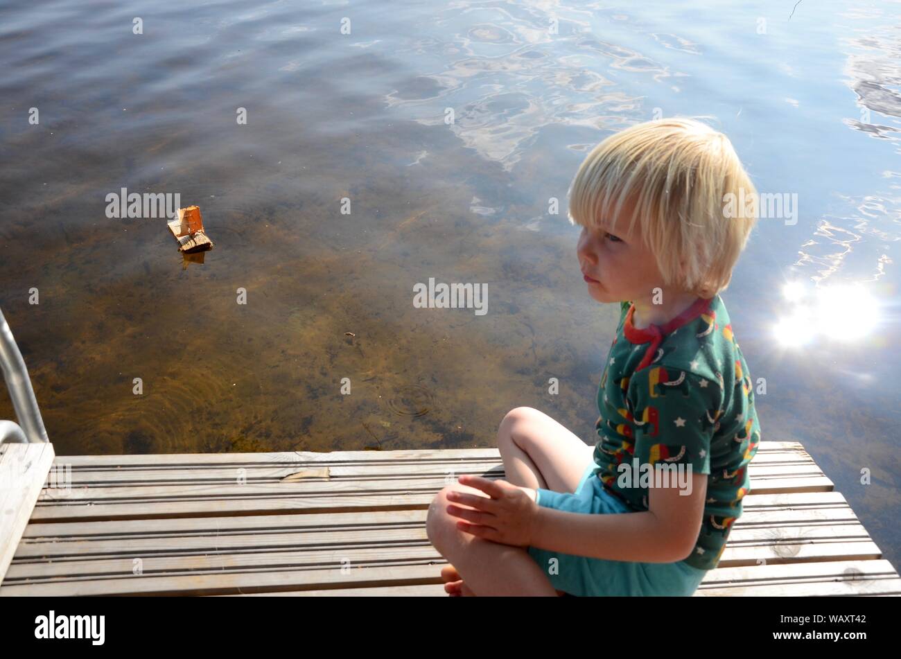 Young blonde boy sitting on jetty with wooden toy boat on lake in ...