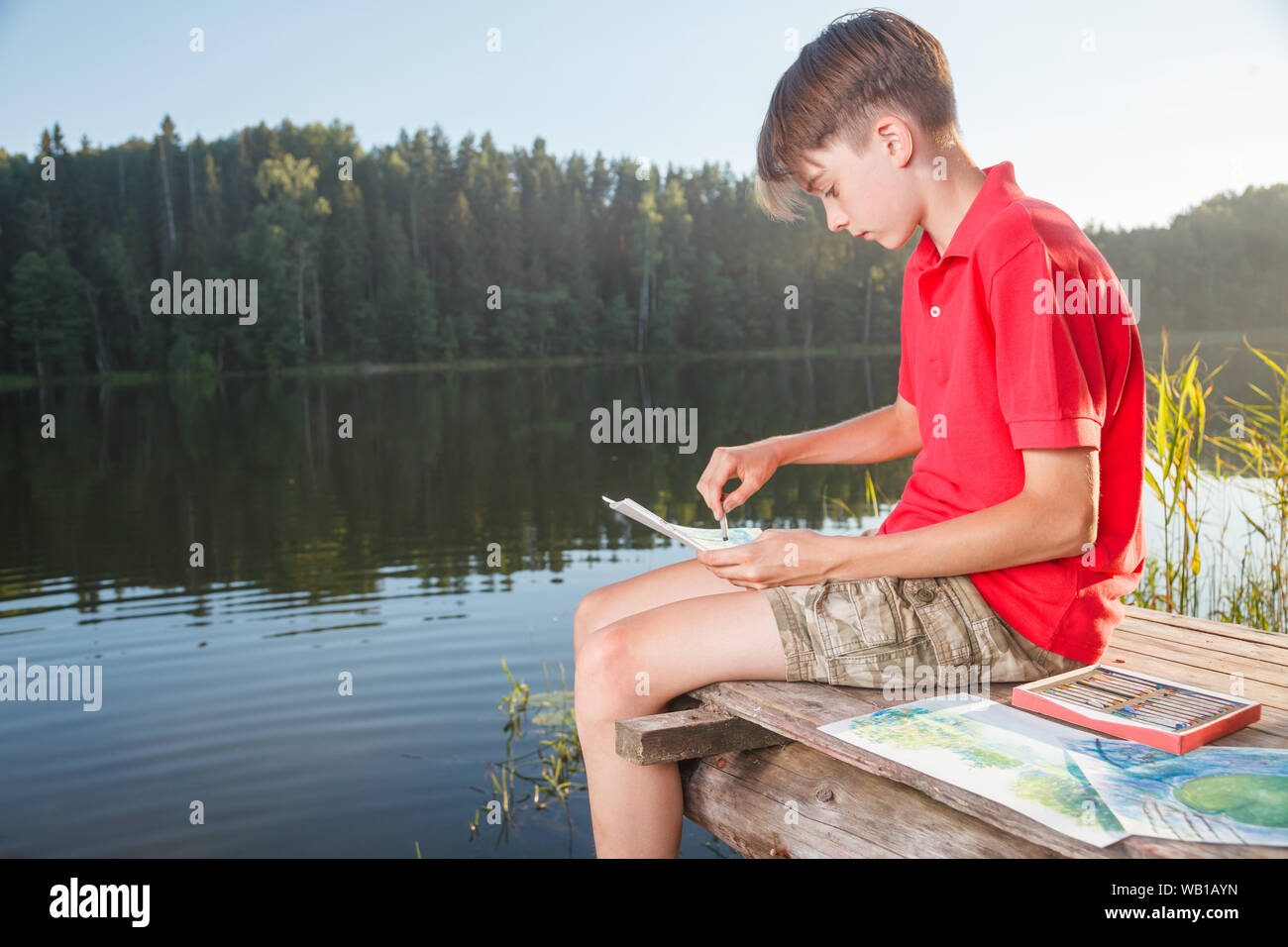 Teenager boy sitting on a wooden pier by forest lake drawing with ...
