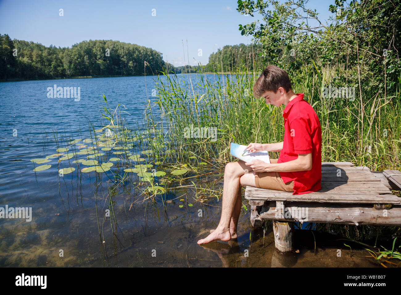 Teenager boy sitting on a wooden pier by forest lake drawing with ...