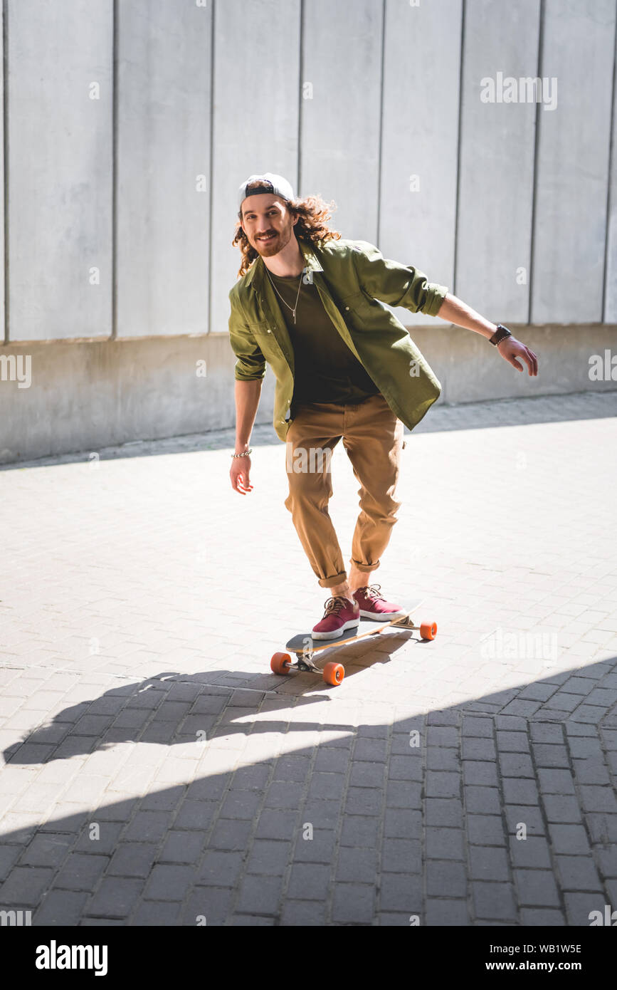 happy man riding on skateboard near concrete wall, looking at camera ...