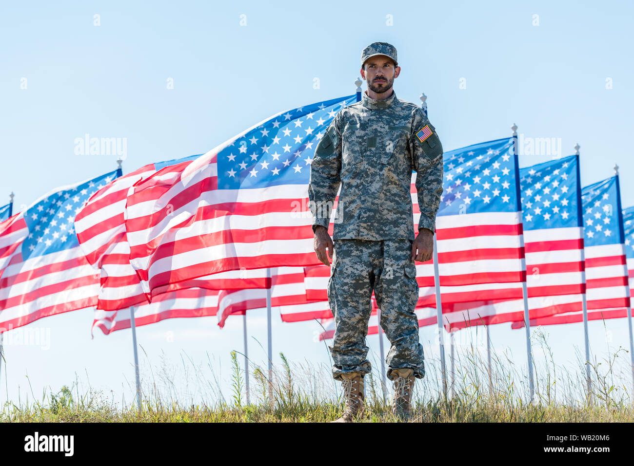 handsome soldier in uniform standing near american flags and blue sky ...