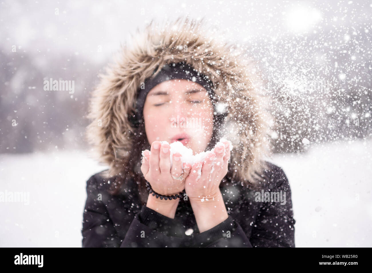 Happy young woman wearing winter clothes while blowing snow on snowy ...