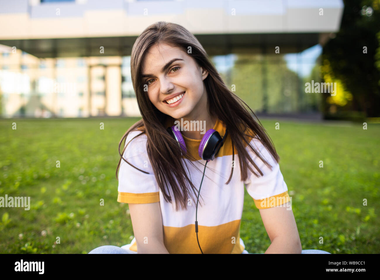 Portrait Of Smiling Cute College Girl With Headphones Posing In Front Of Her Alma Mater Stock