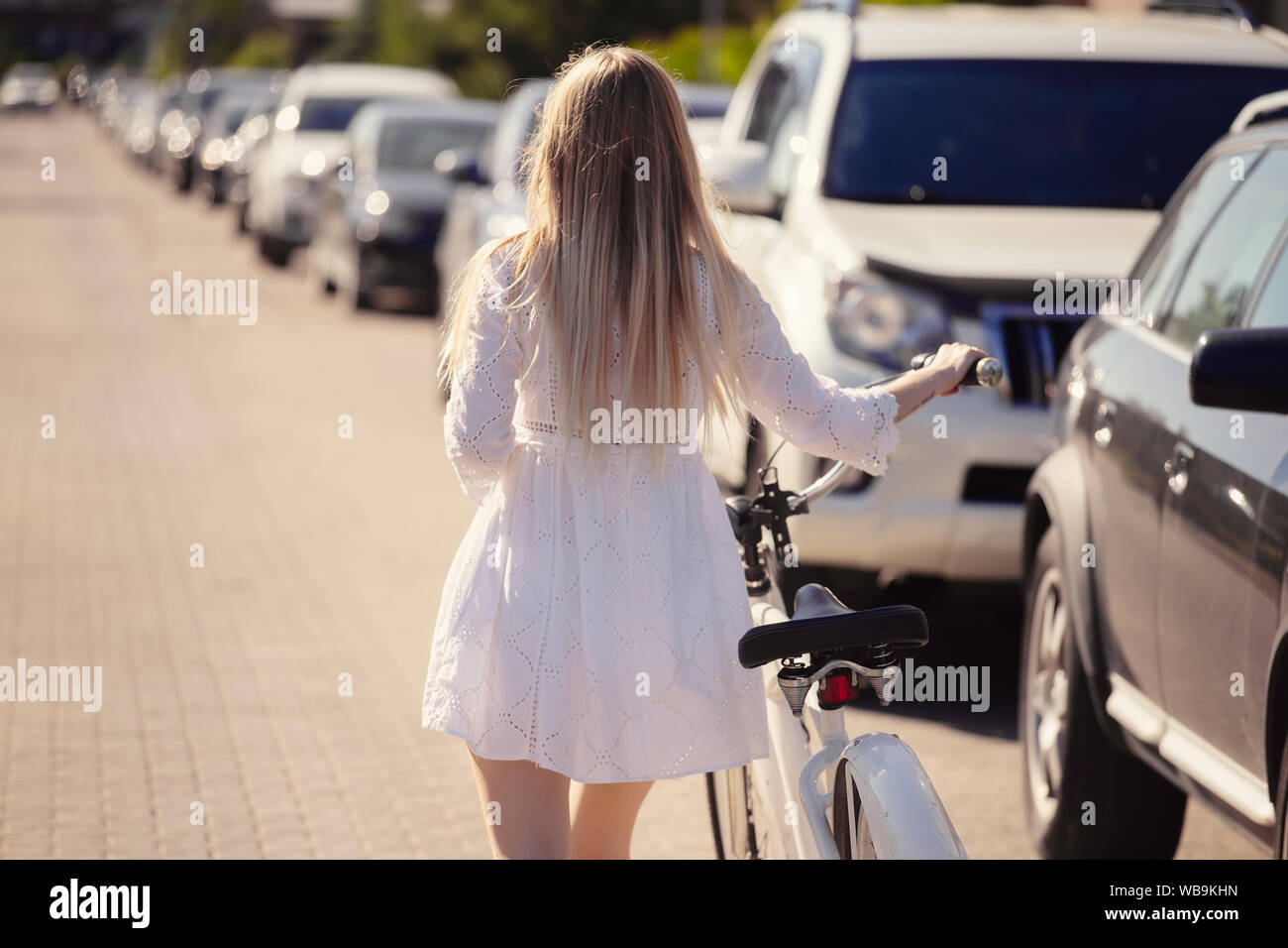 young blond woman in white dress walking with bike near cars at city, rear view Stock Photo - Alamy