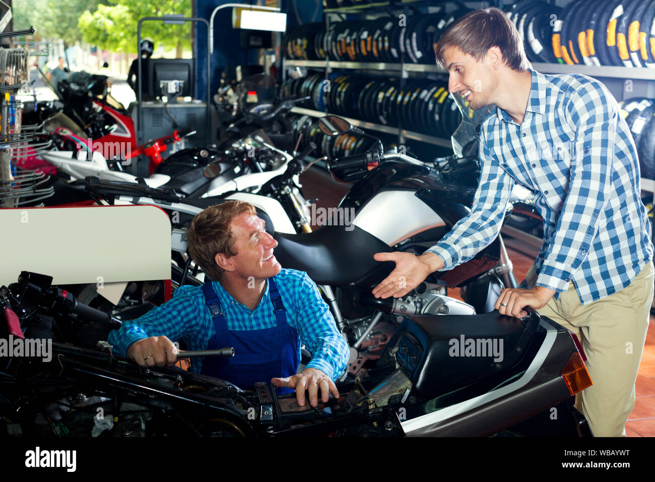 Adult male technician standing with customer in shop with motorcycles ...