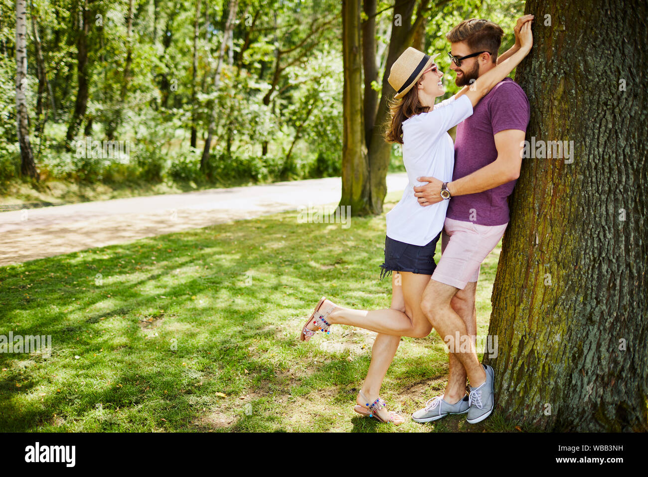 Young Couple Embracing In The Park Leaning Against A Tree And Looking