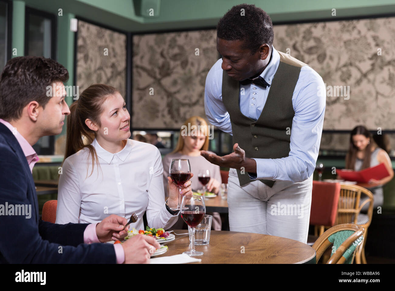 Positive elegant couple talking with polite African American waiter ...