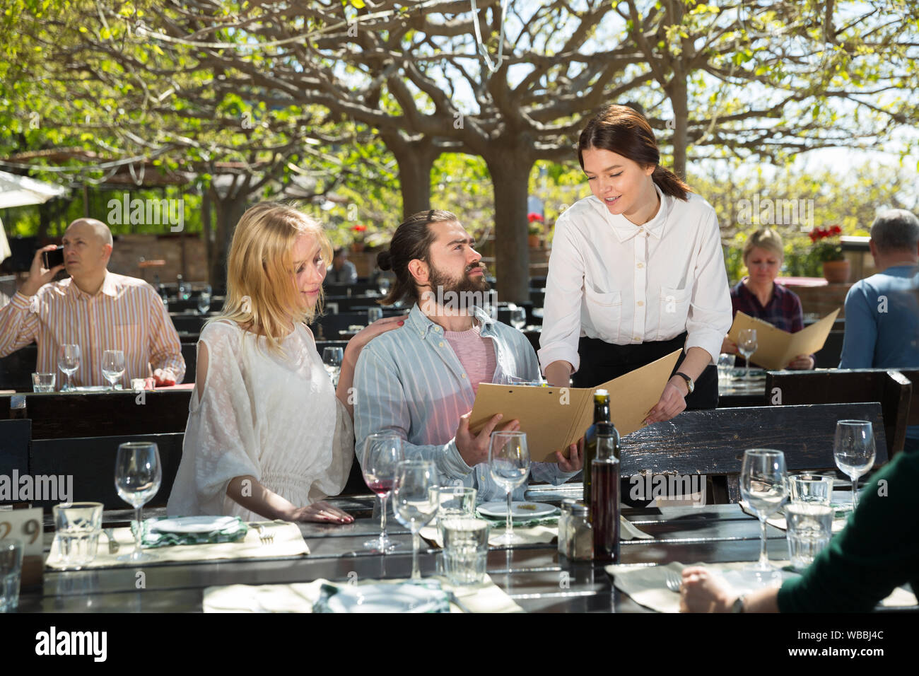 Polite young waitress showing menu card to smiling couple, recommending ...