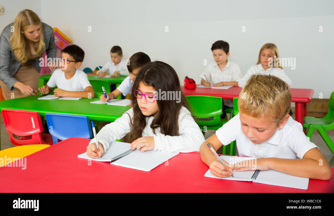 Group of school kids with pens and notebooks studying in classroom with ...