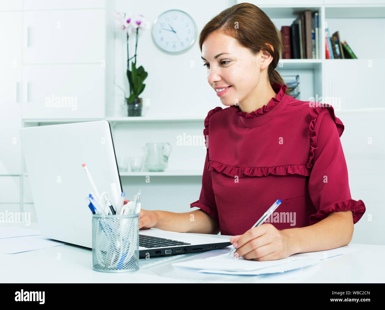 Young woman employee taking notes on paper while working with laptop at ...