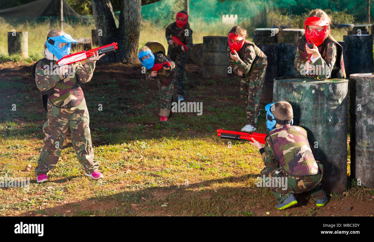 Group of smiling kids in camouflages and masks playing paintball aiming ...