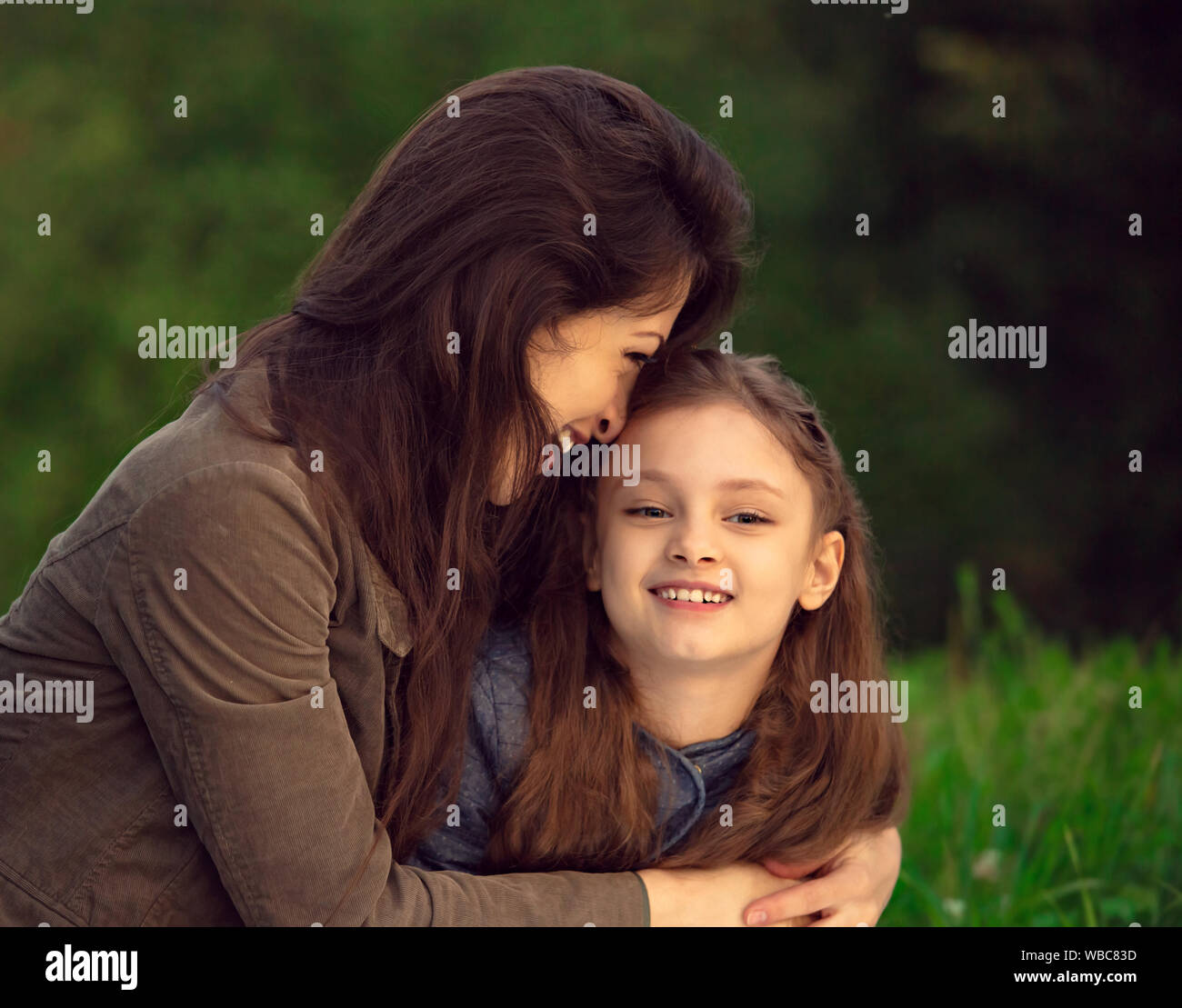 Beautiful young laughing mother embracing her cute long hair daughter on summer green grass ...