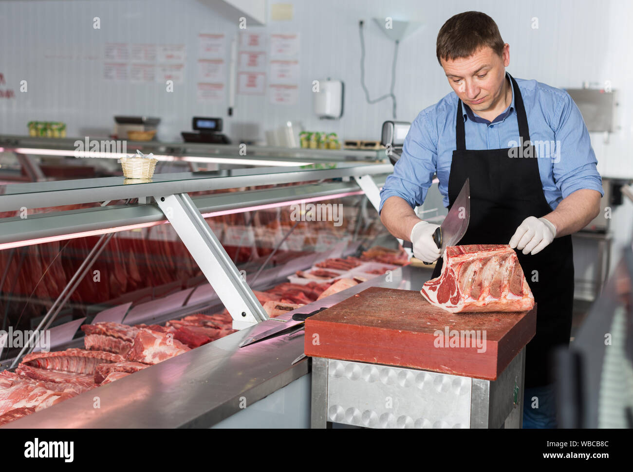 Confident seller of butcher store cutting fresh raw steak on ribs with ...