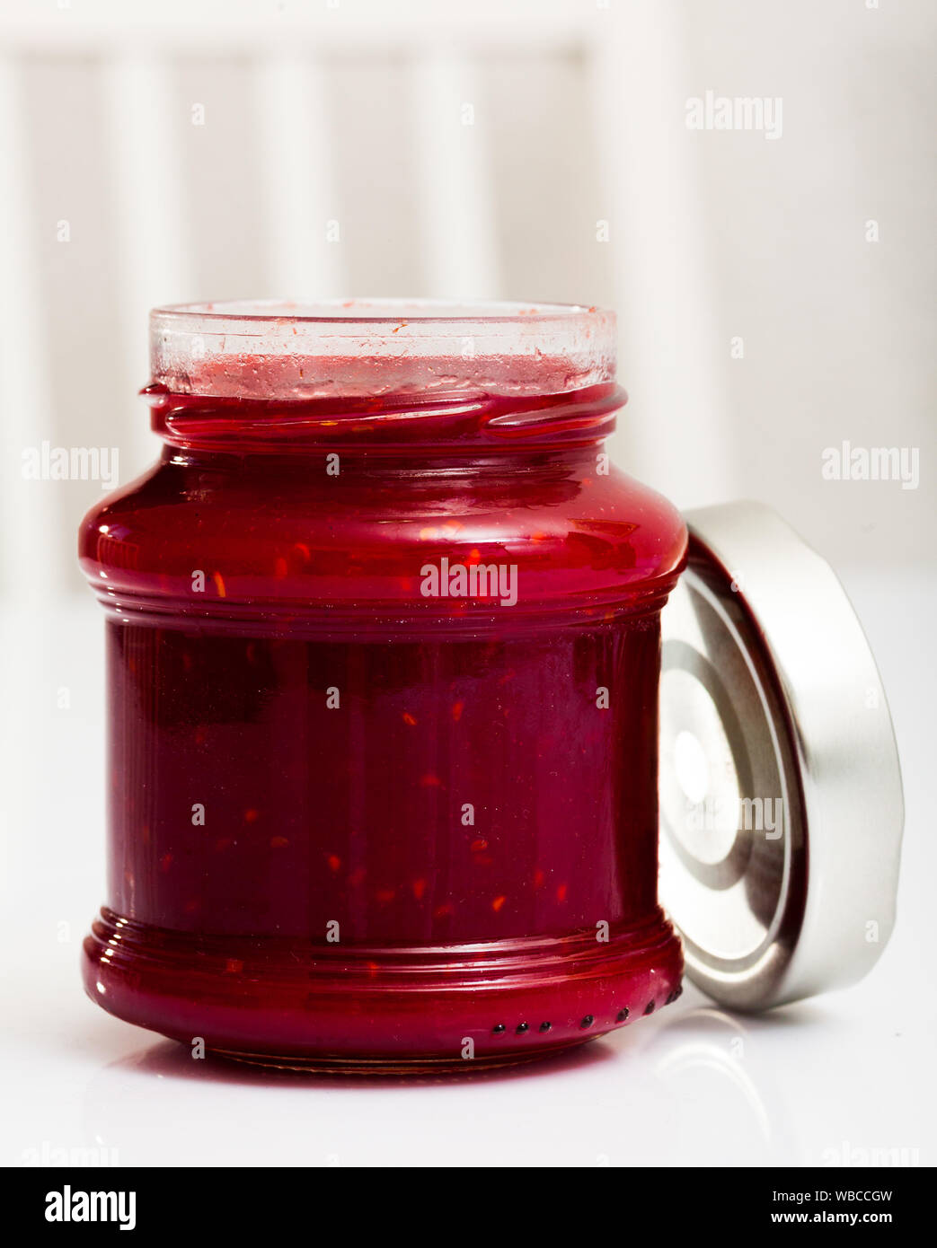 Open glass jar of delicious homemade raspberry jam on white surface ...