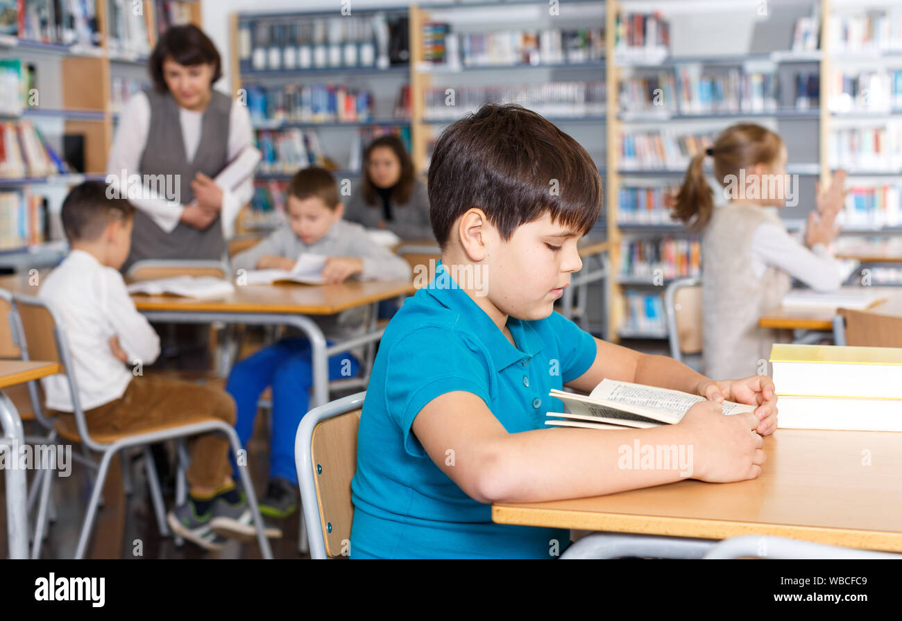 Focused boy reading in school library on background with other students ...