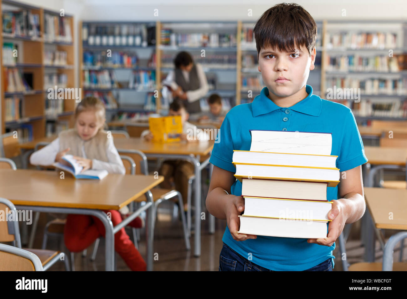 Upset tween boy standing in school library with pile of books in hands ...