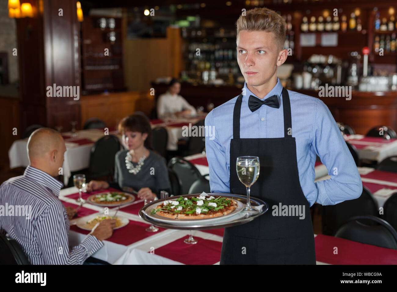 Portrait of smiling waiter with serving tray meeting restaurant guests ...