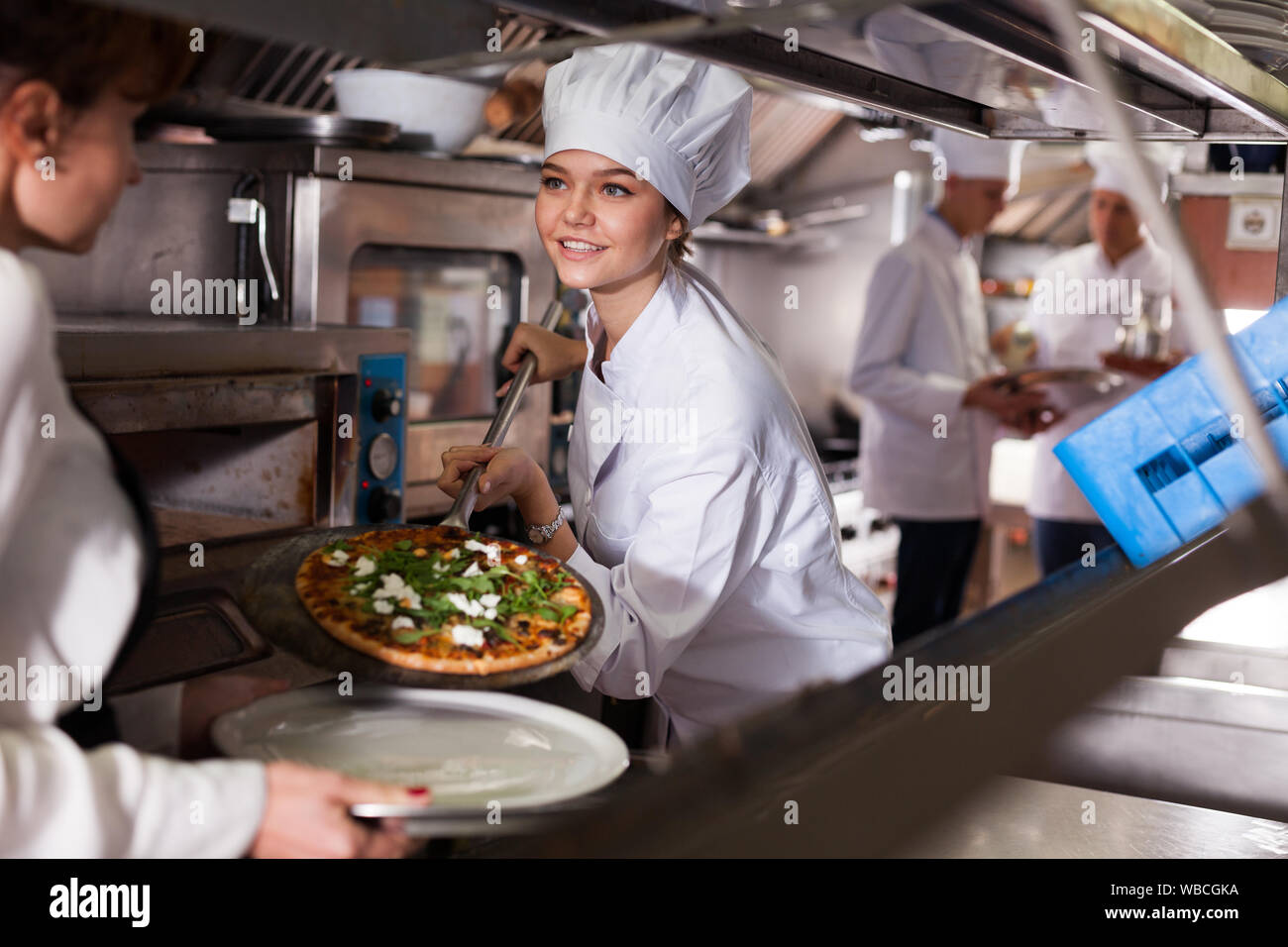 Smiling young female chef giving cooked pizza to waitress ready to ...