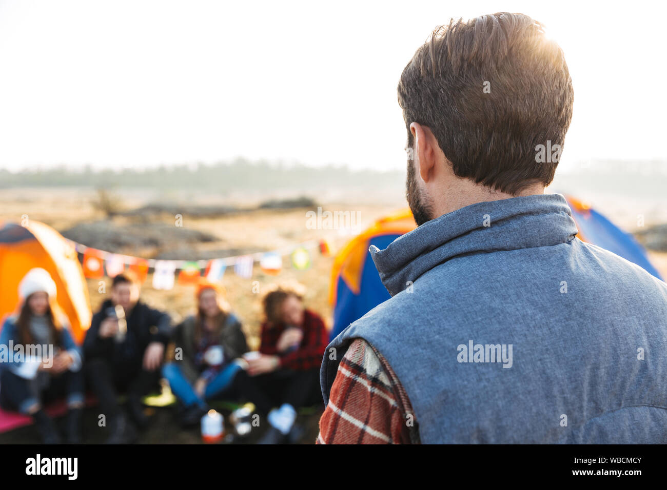Back view of a young man standing at the campsite with his friends ...