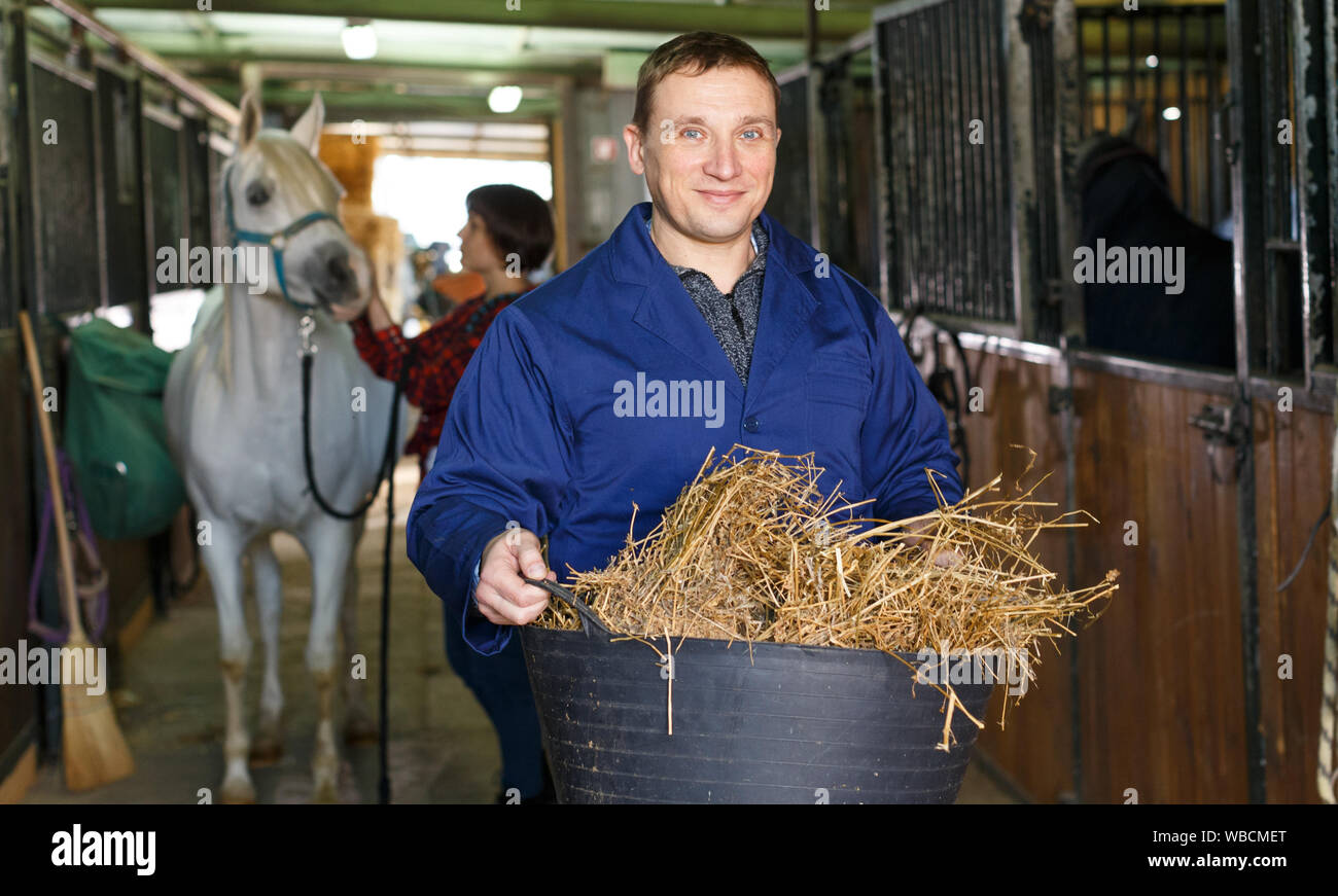 Man in working clothes feeding horse with hay at stable Stock Photo - Alamy