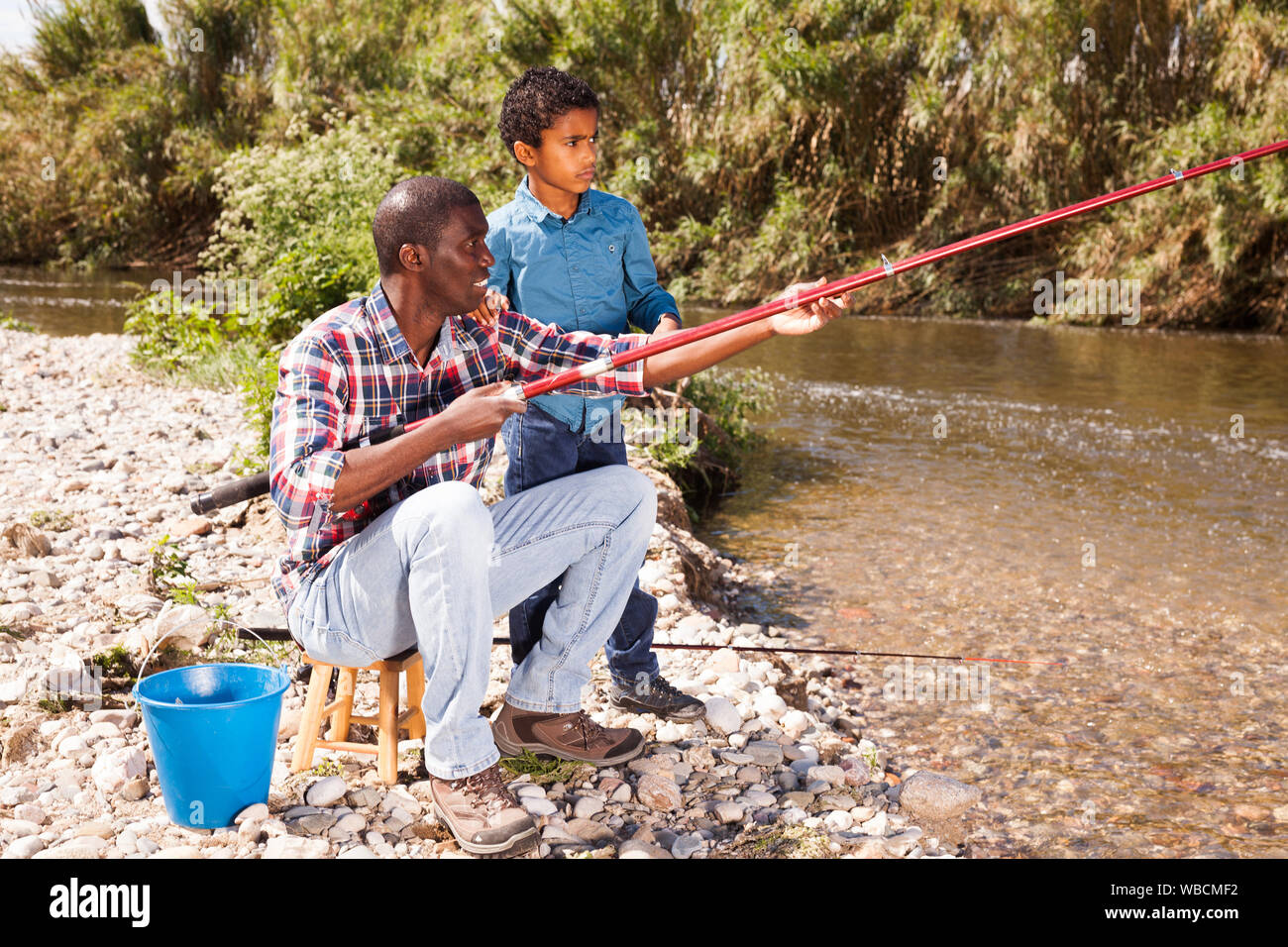 Portrait of cheerful afro man and his little son fishing with rods on ...