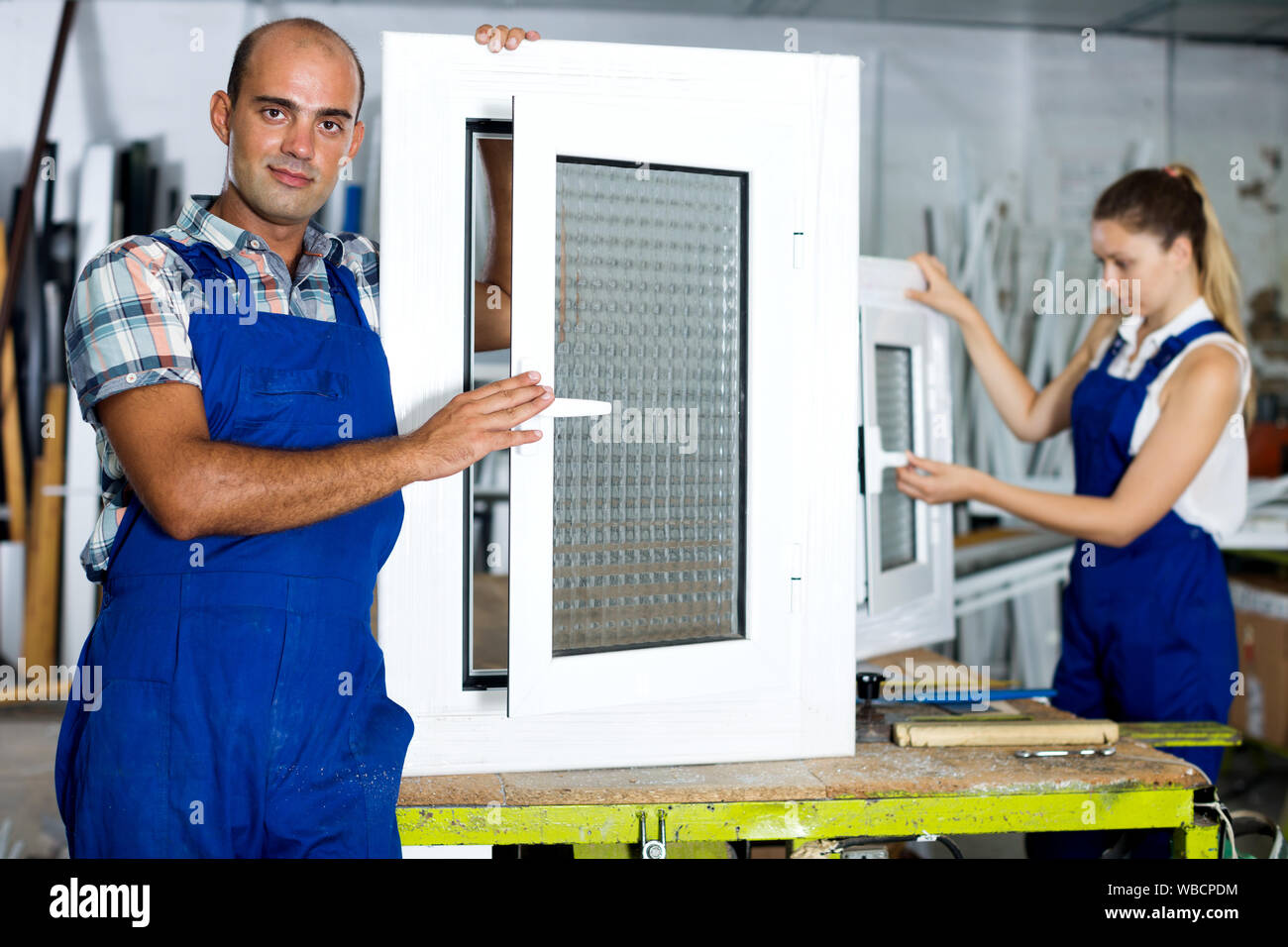 Positive workman in blue overalls demonstrating pvc window in workshop ...