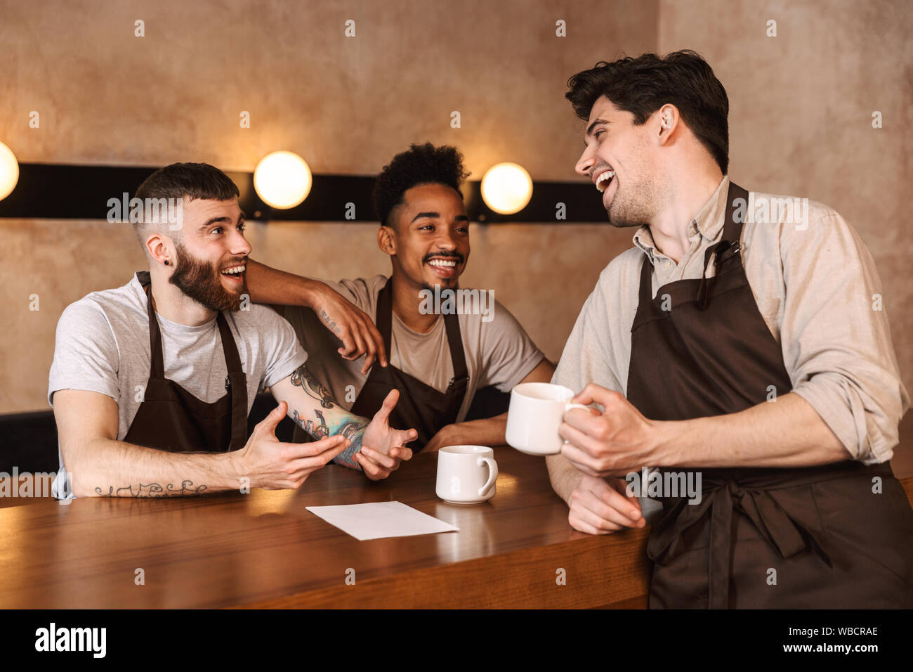 Three cheerful men baristas at the coffee shop, drinking coffee during ...
