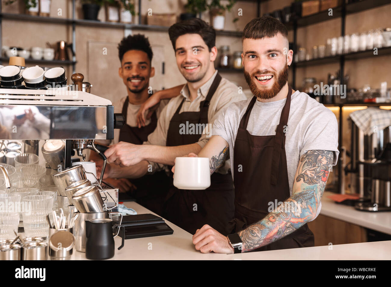 Three cheerful male baristas standing at the coffee shop counter ...