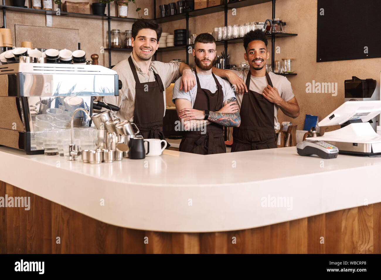 Three cheerful male baristas standing at the coffee shop counter ...