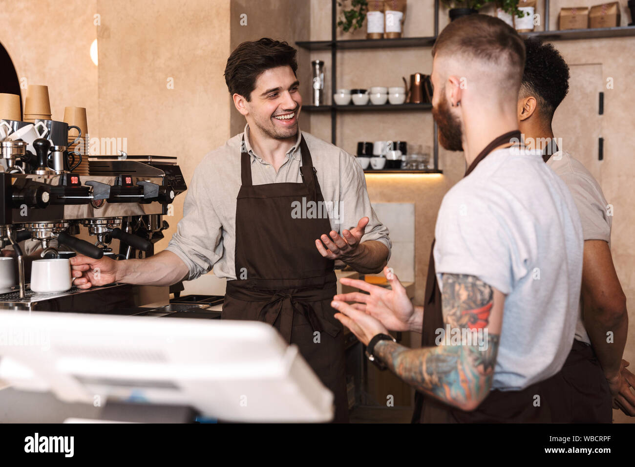 Three cheerful male baristas standing at the coffee shop counter ...