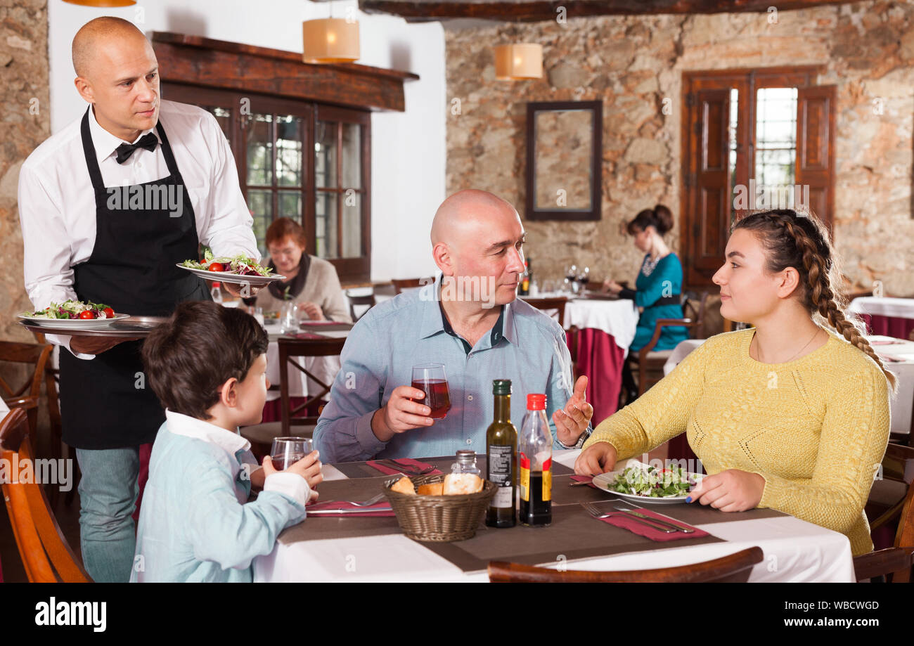Cheerful waiter serving plates of salad to happy family having dinner ...