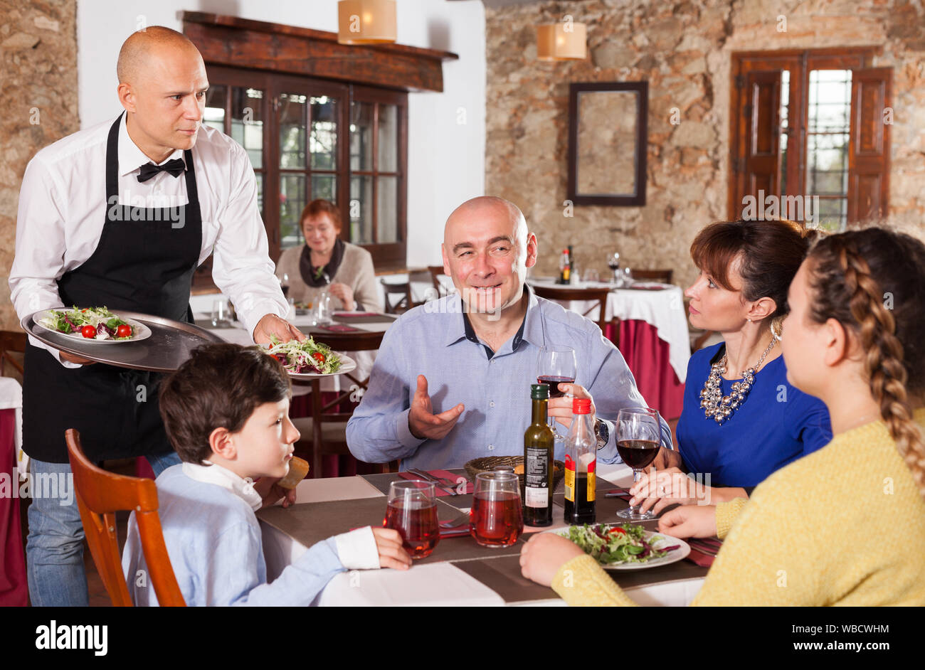 Cheerful waiter serving plates of salad to happy family having dinner ...