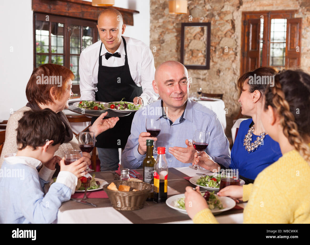 Cheerful waiter serving plates of salad to happy family having dinner ...