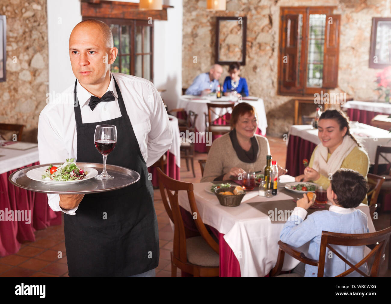 Positive waiter with tray in foreground and visitors having dinner at ...