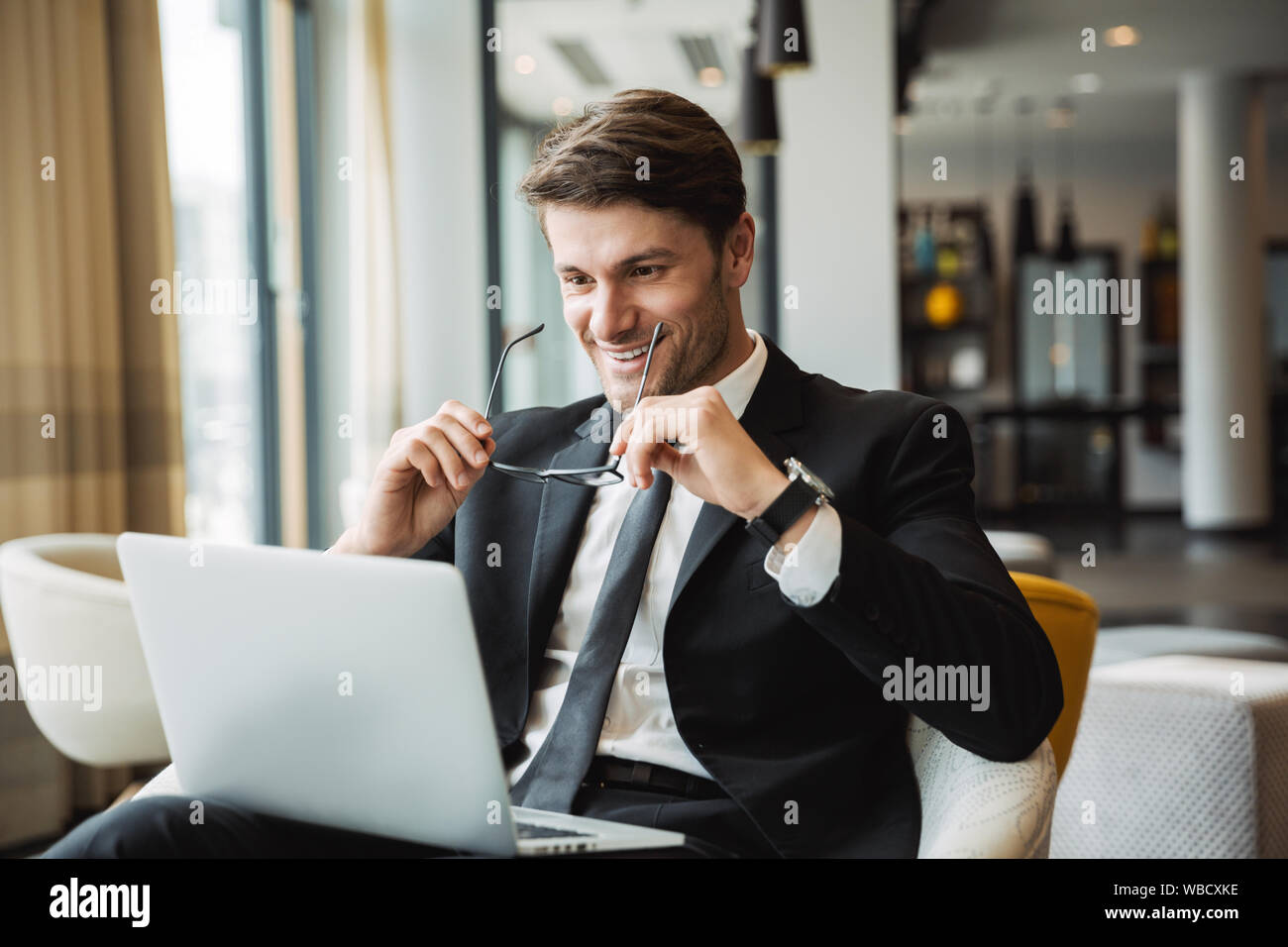 Portrait of smiling young businessman wearing formal black suit sitting on armchair with laptop ...