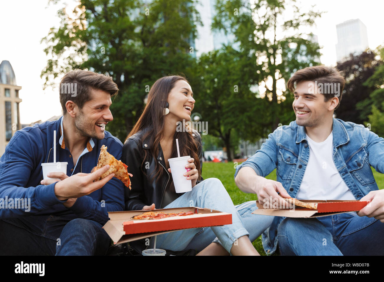 Group of happy young friends eating pizza while sitting on a grass at ...