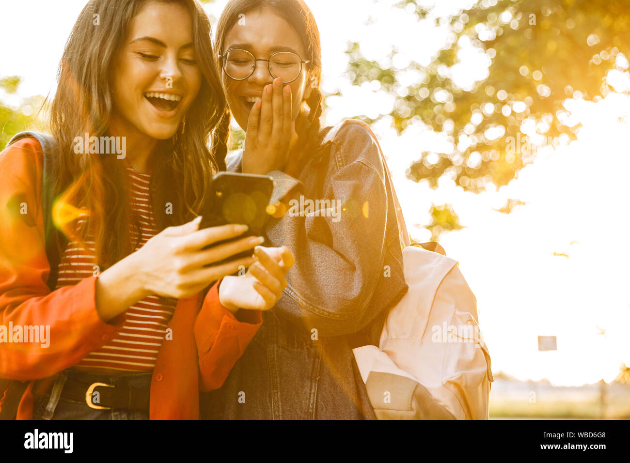 Image of two delighted girls wearing backpacks laughing and typing on ...