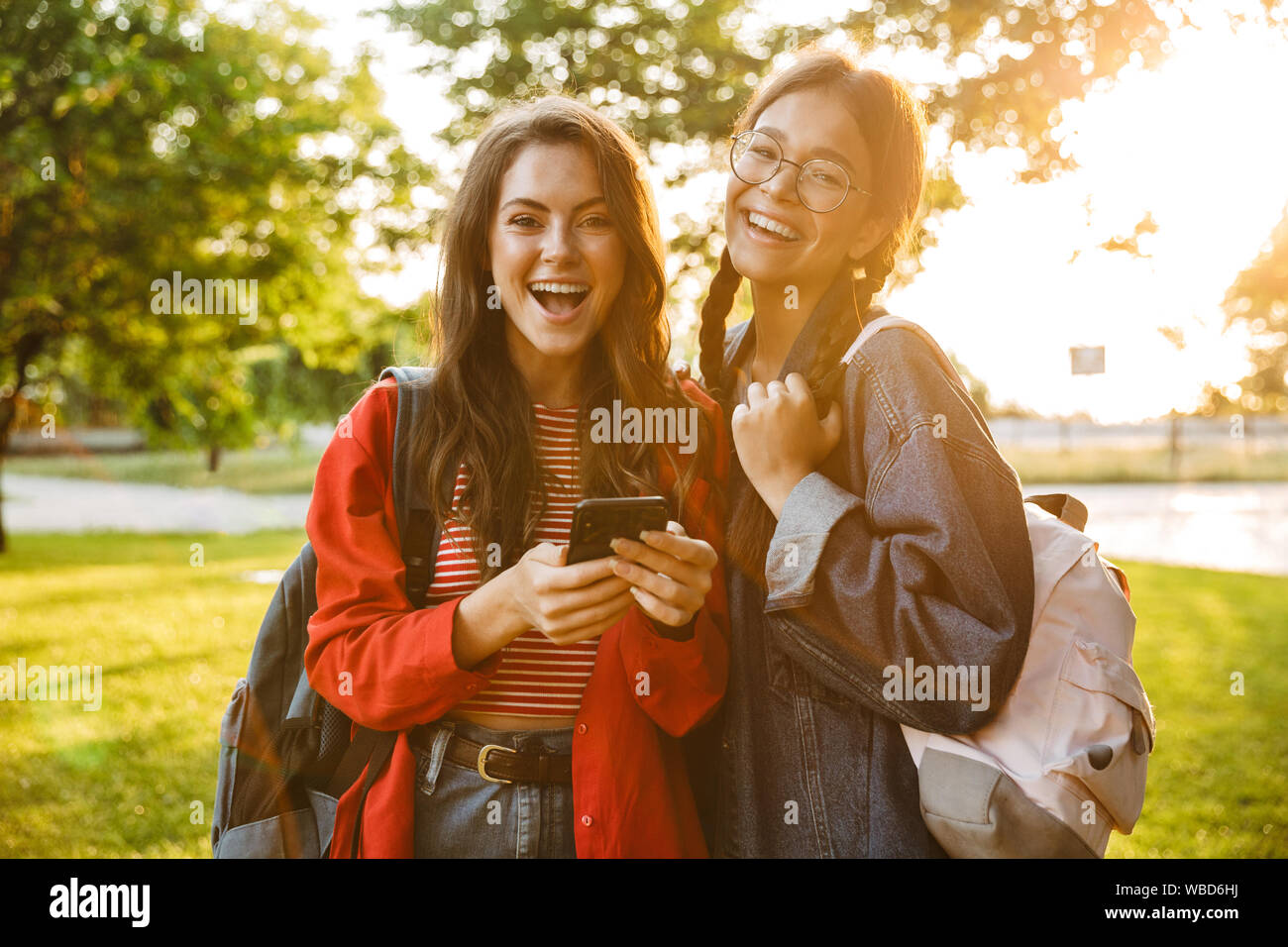 Image of two delighted girls wearing backpacks smiling and typing on ...
