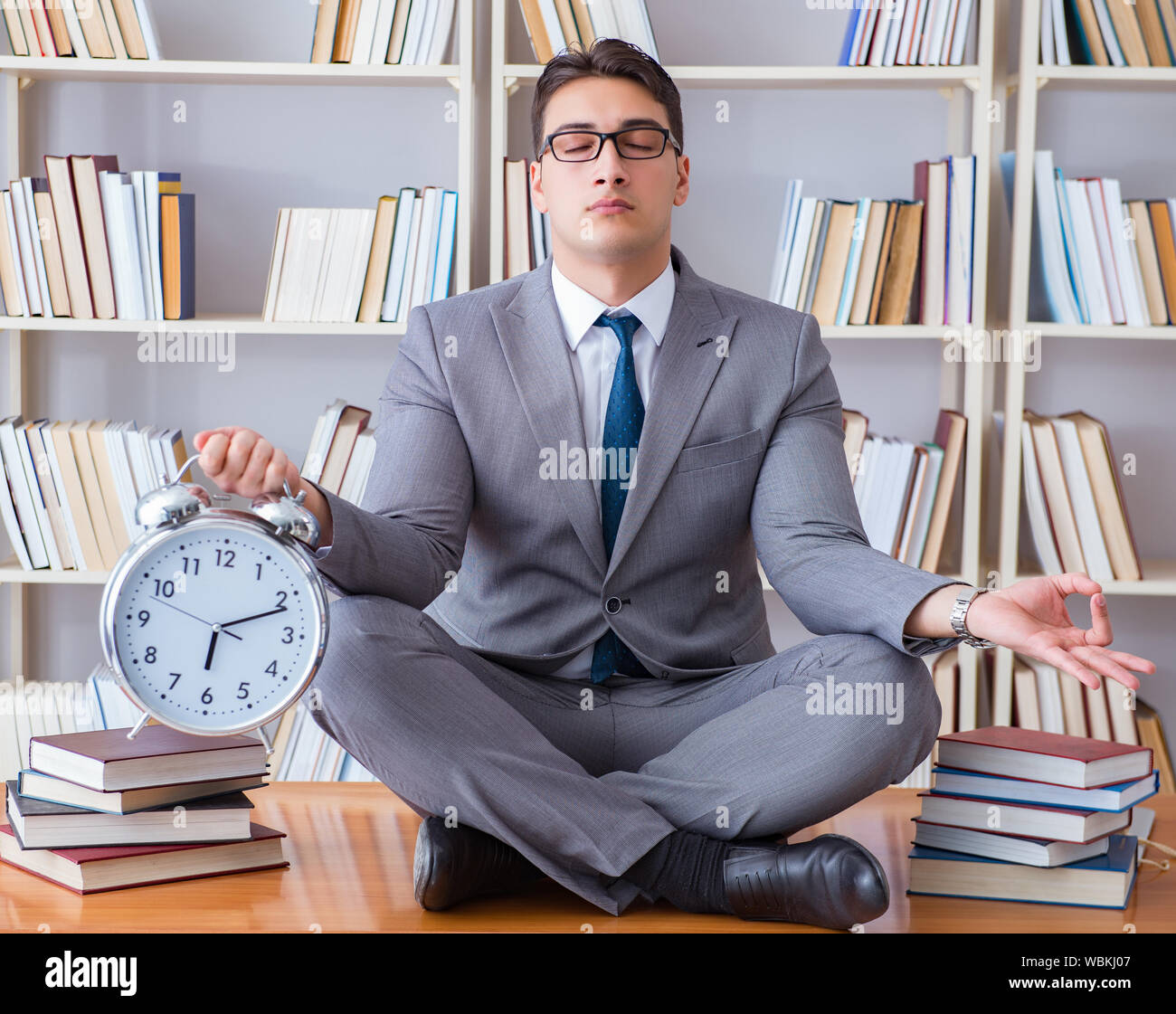Businessman student in lotus position with an alarm clock in library ...