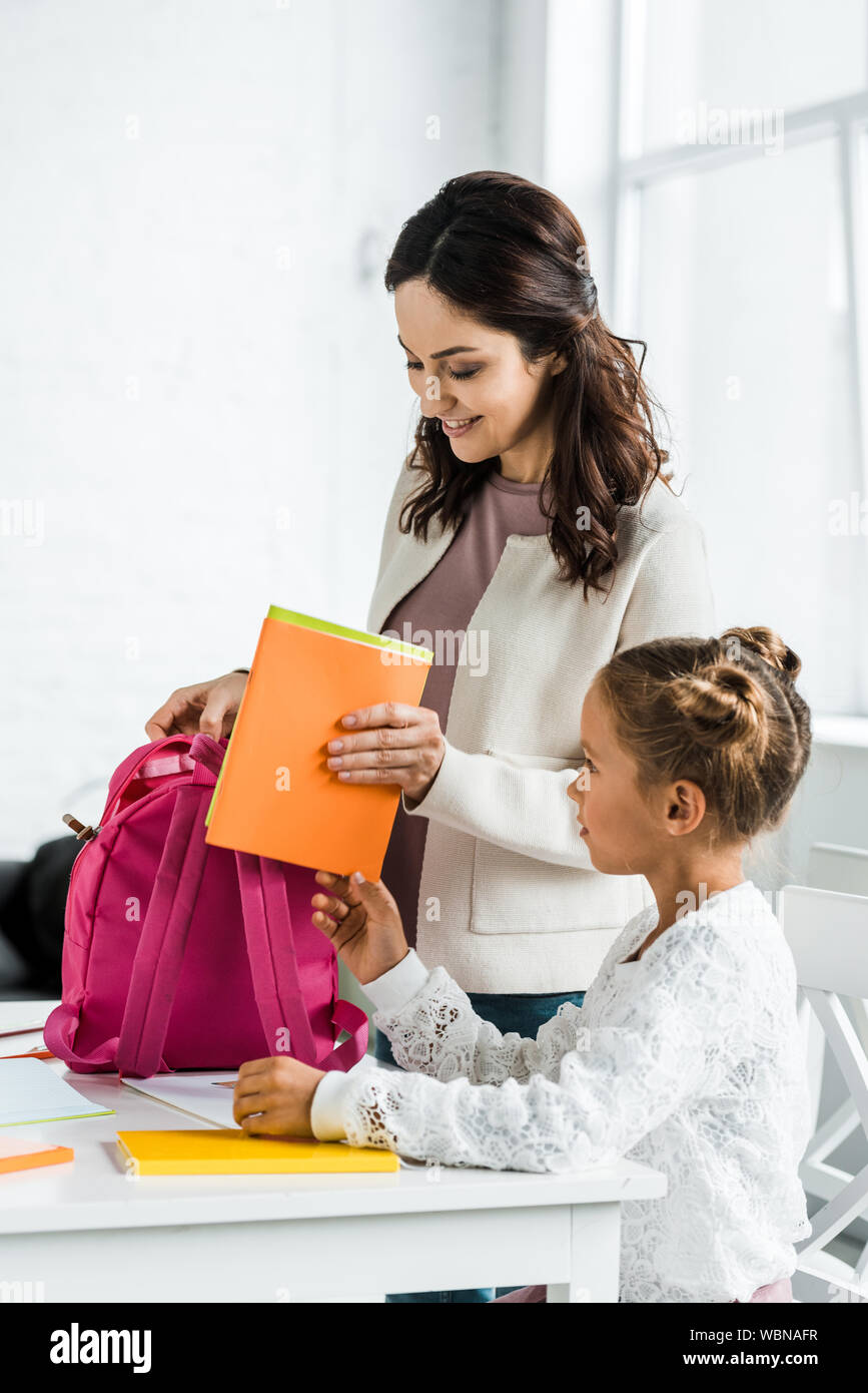 happy mother putting book in backpack of schoolgirl at home Stock Photo ...