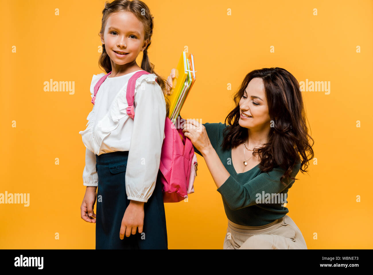 cheerful mother putting books in backpack of schoolgirl isolated on ...