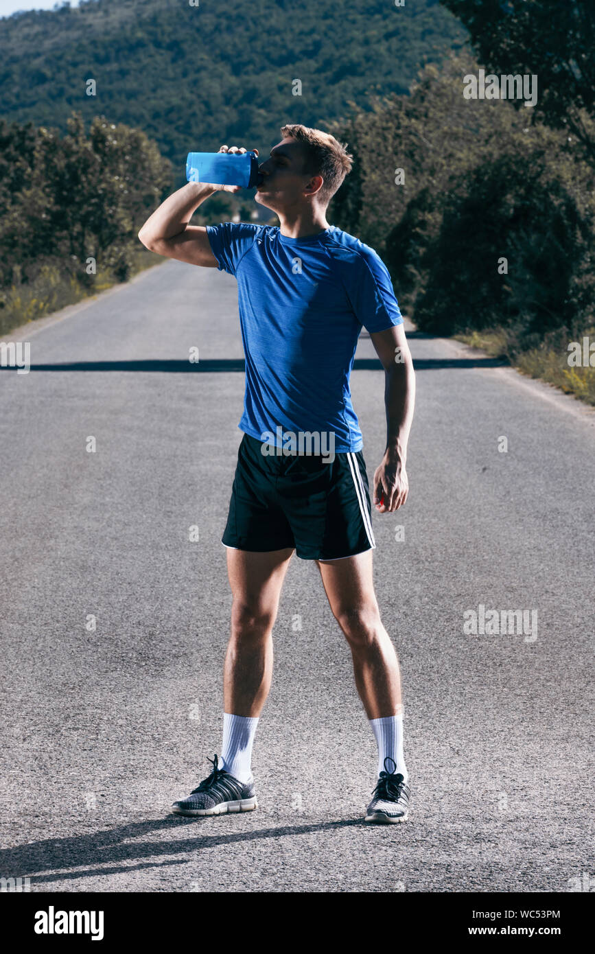 Fit male runner drinking water from a water bottle on an empty road in ...