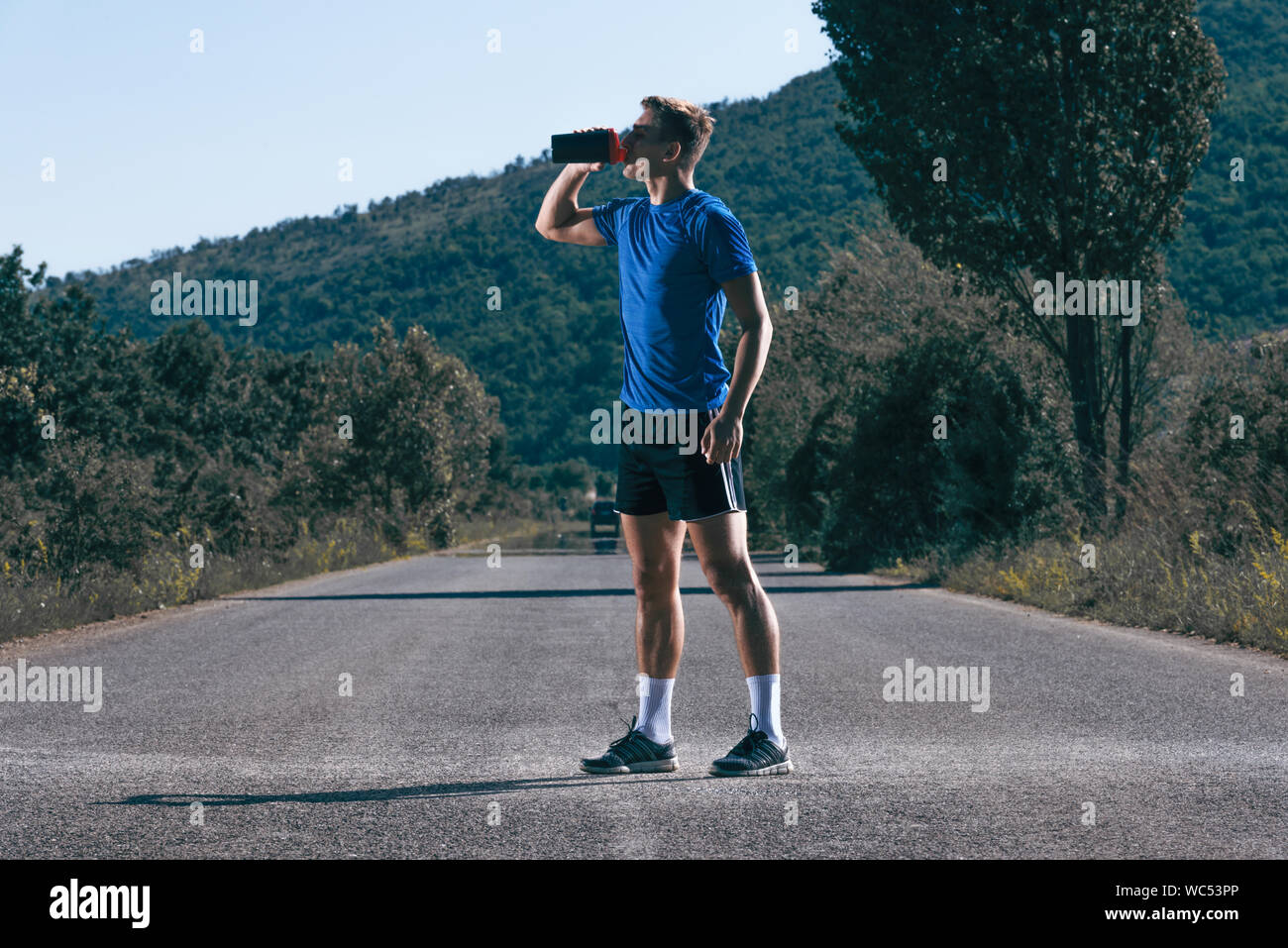 Fit male runner drinking water from a water bottle on an empty road in ...