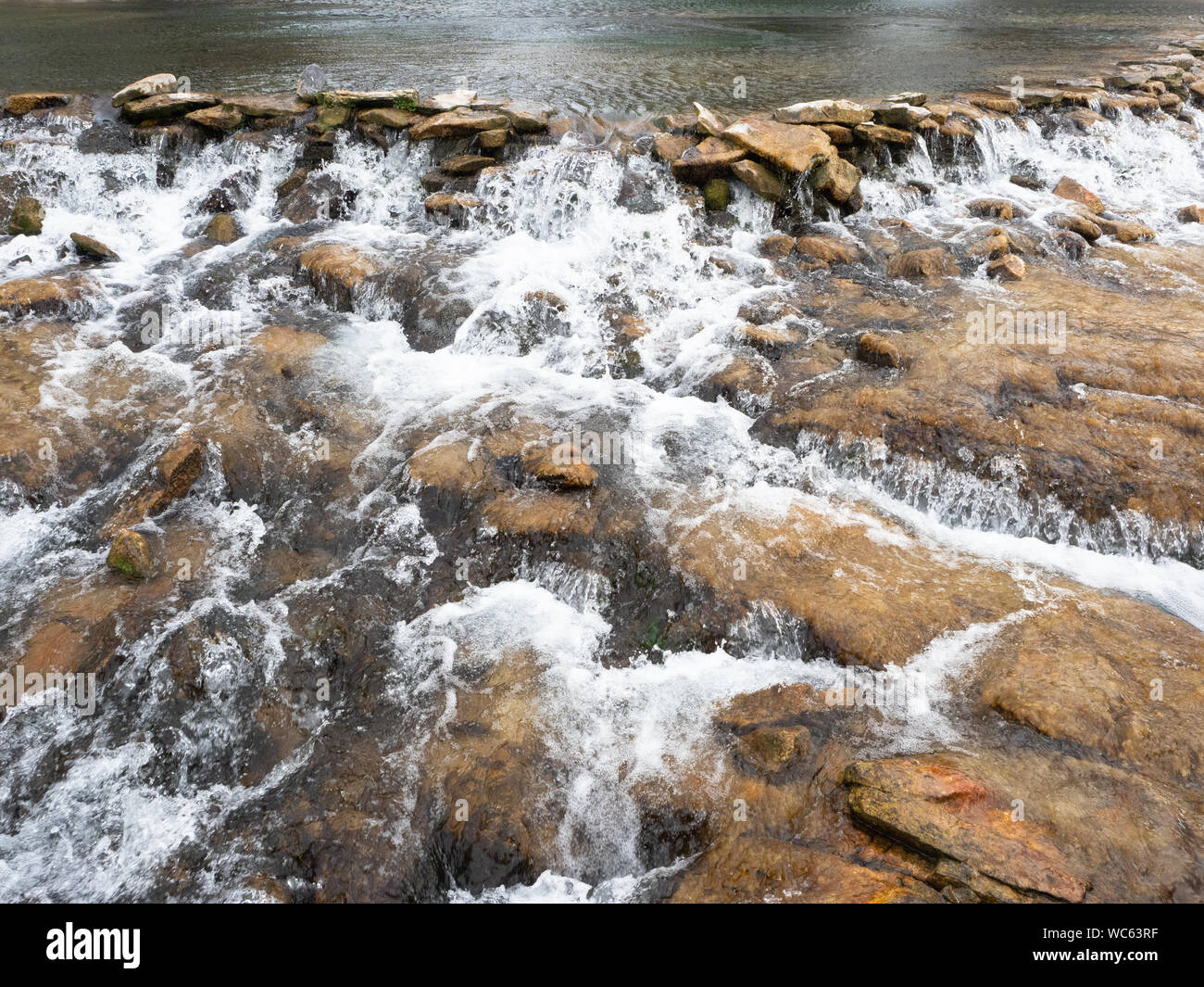 Water falling over brown and rust-colored rocks with a river in the ...