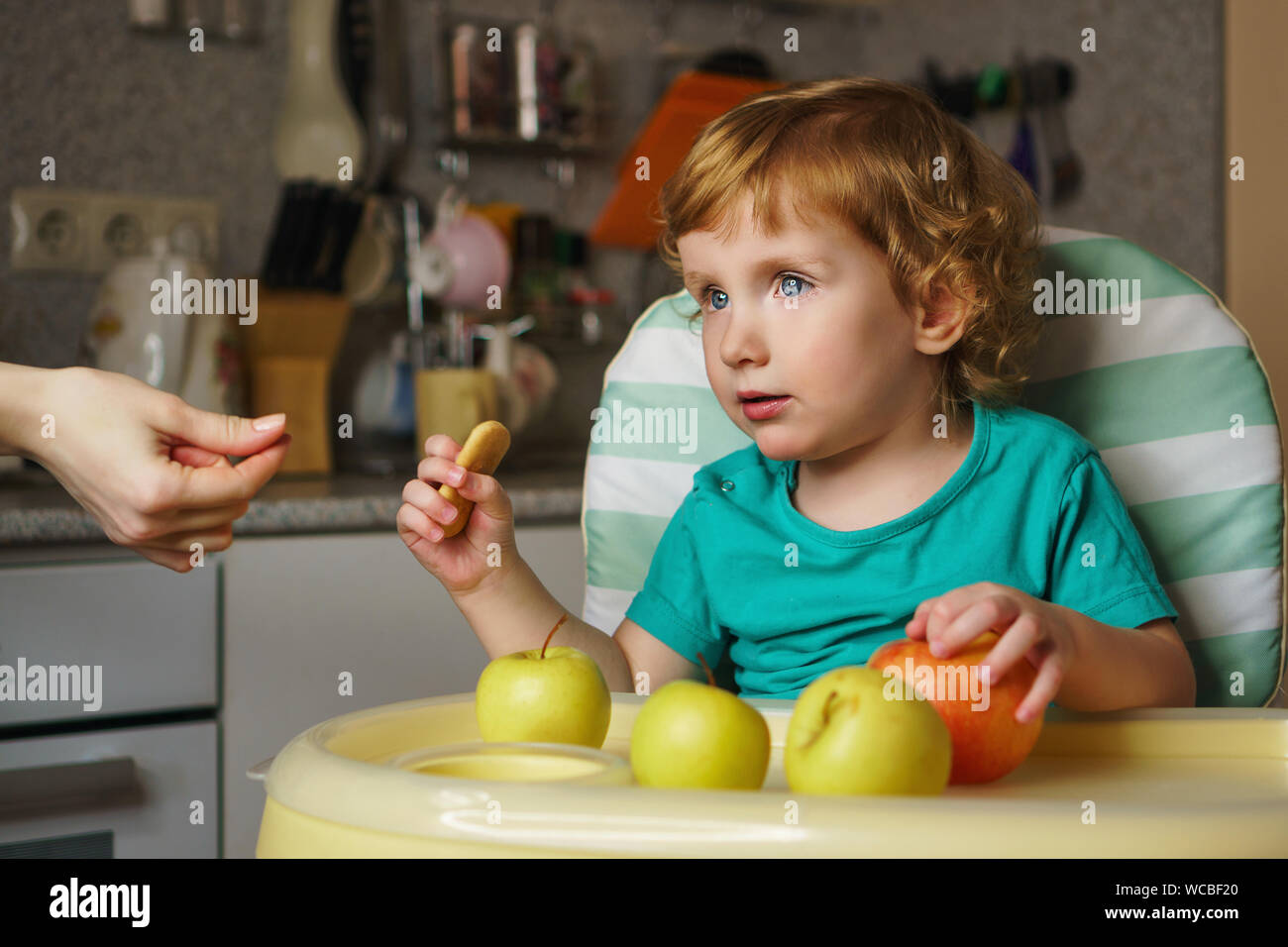 Charming little child eating in the kitchen. He sits in a highchair ...