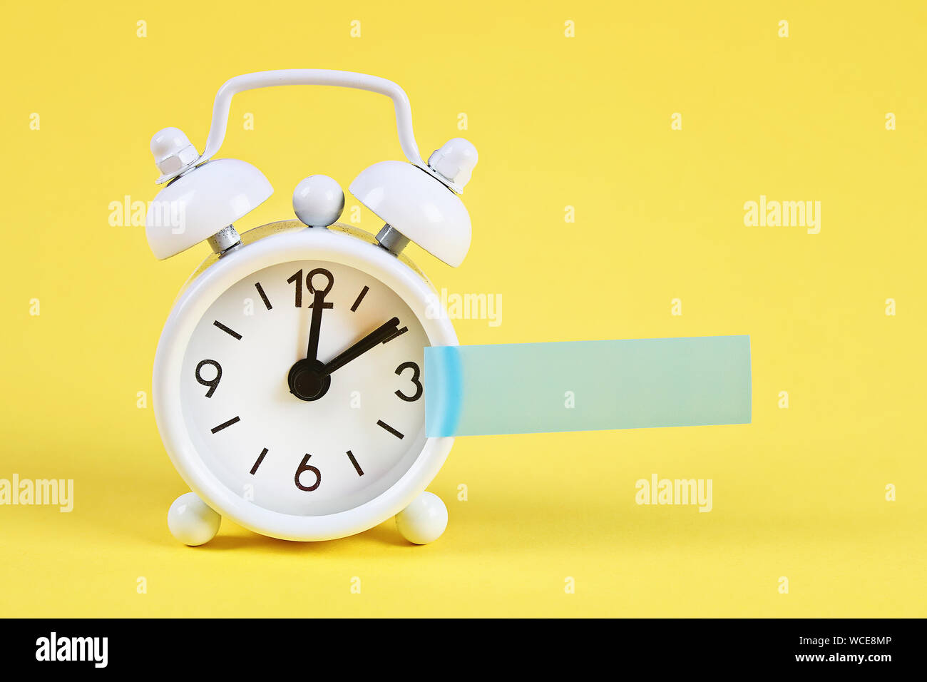 White alarm clock on yellow pastel background. Blank sticky note on the ...