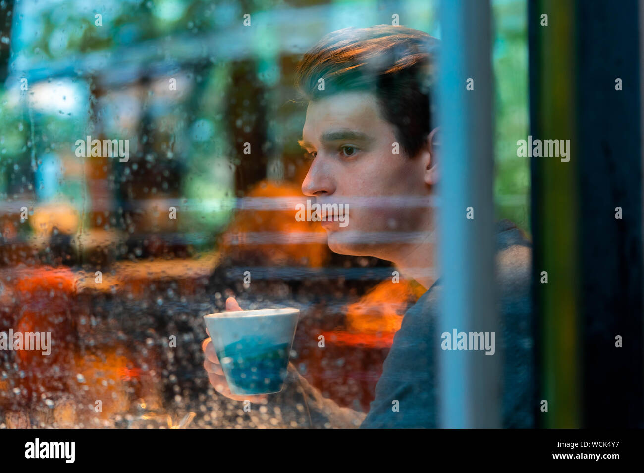 Young pensive handsome man drinking tea or coffee on rainy day in cozy ...