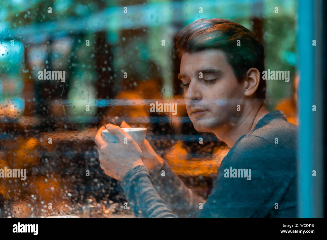 Young pensive handsome man drinking tea or coffee on rainy day in cozy ...